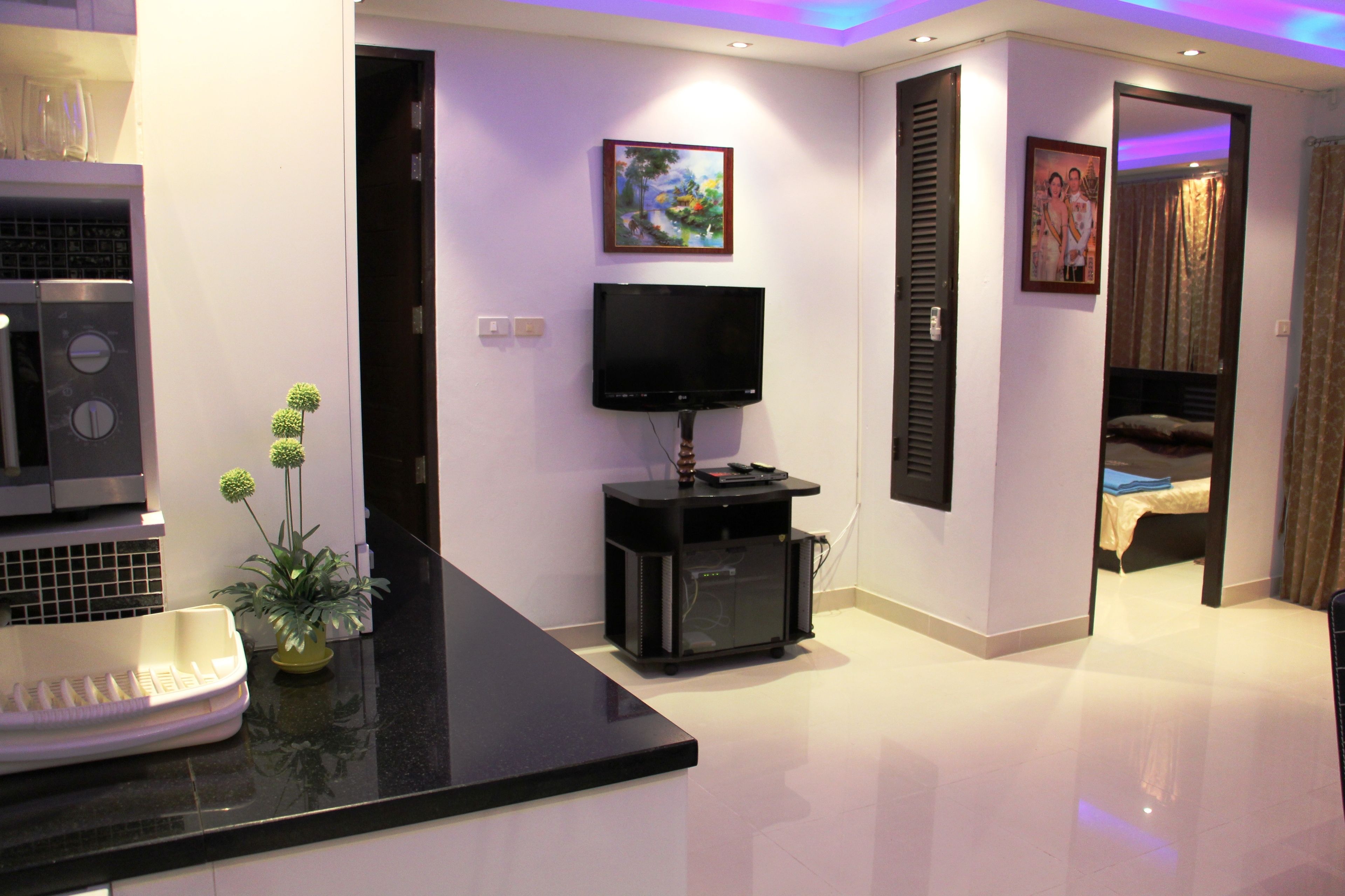 Uma sala de estar moderna com uma TV de tela plana, um móvel de entretenimento preto pequeno, uma área de jantar com uma mesa e cadeiras brancas, e uma cozinha com micro-ondas e pia.