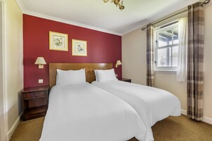 Quarto de hotel com duas camas, uma parede de destaque vermelha, duas imagens emolduradas, abajures, uma janela com cortinas e um candelabro.