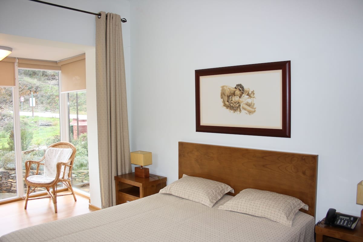 Quarto com cama, cadeira, mesa de cabeceira, abajur e um quadro na parede.