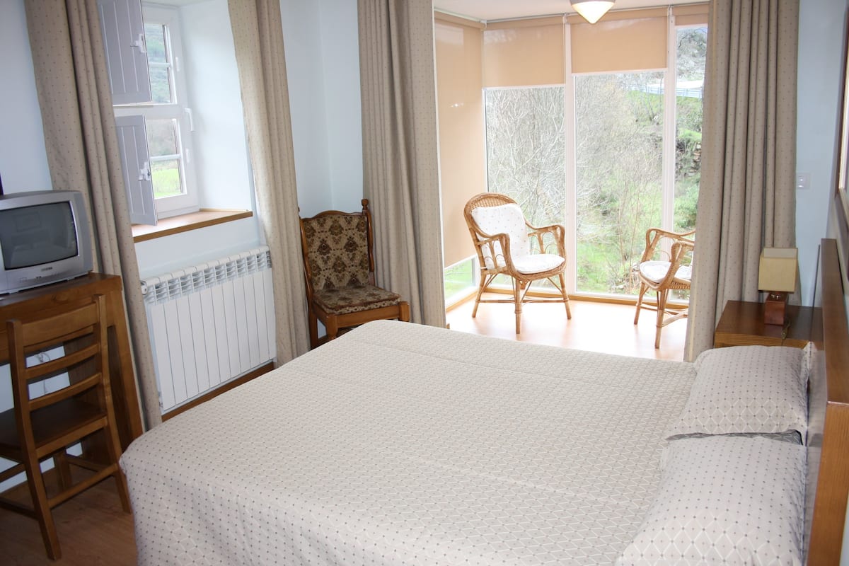 Quarto com uma cama grande, televisão sobre um suporte de madeira, uma cadeira e vista para o exterior pela janela.