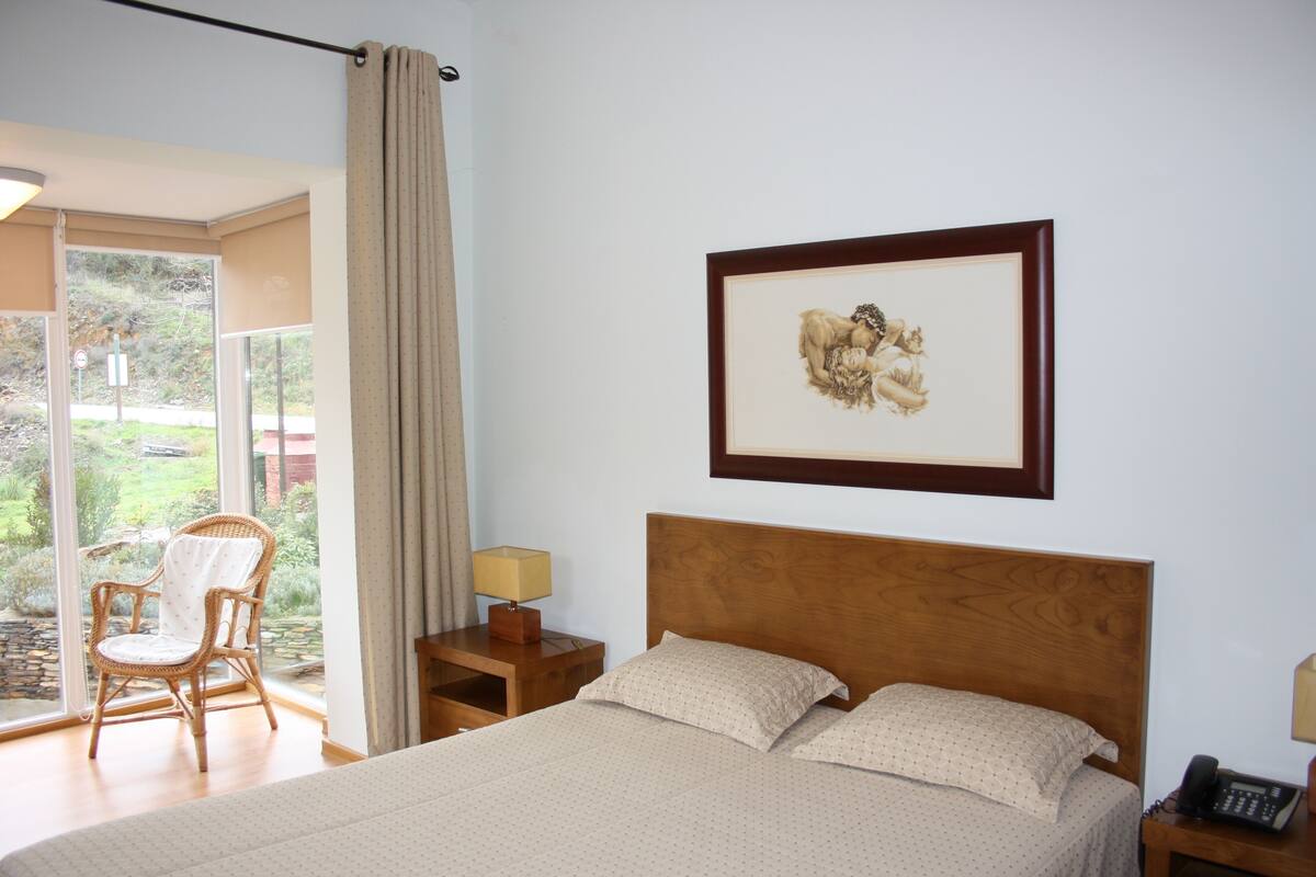 Quarto com cama, cadeira, mesa de cabeceira, telefone e quadro pendurado na parede.