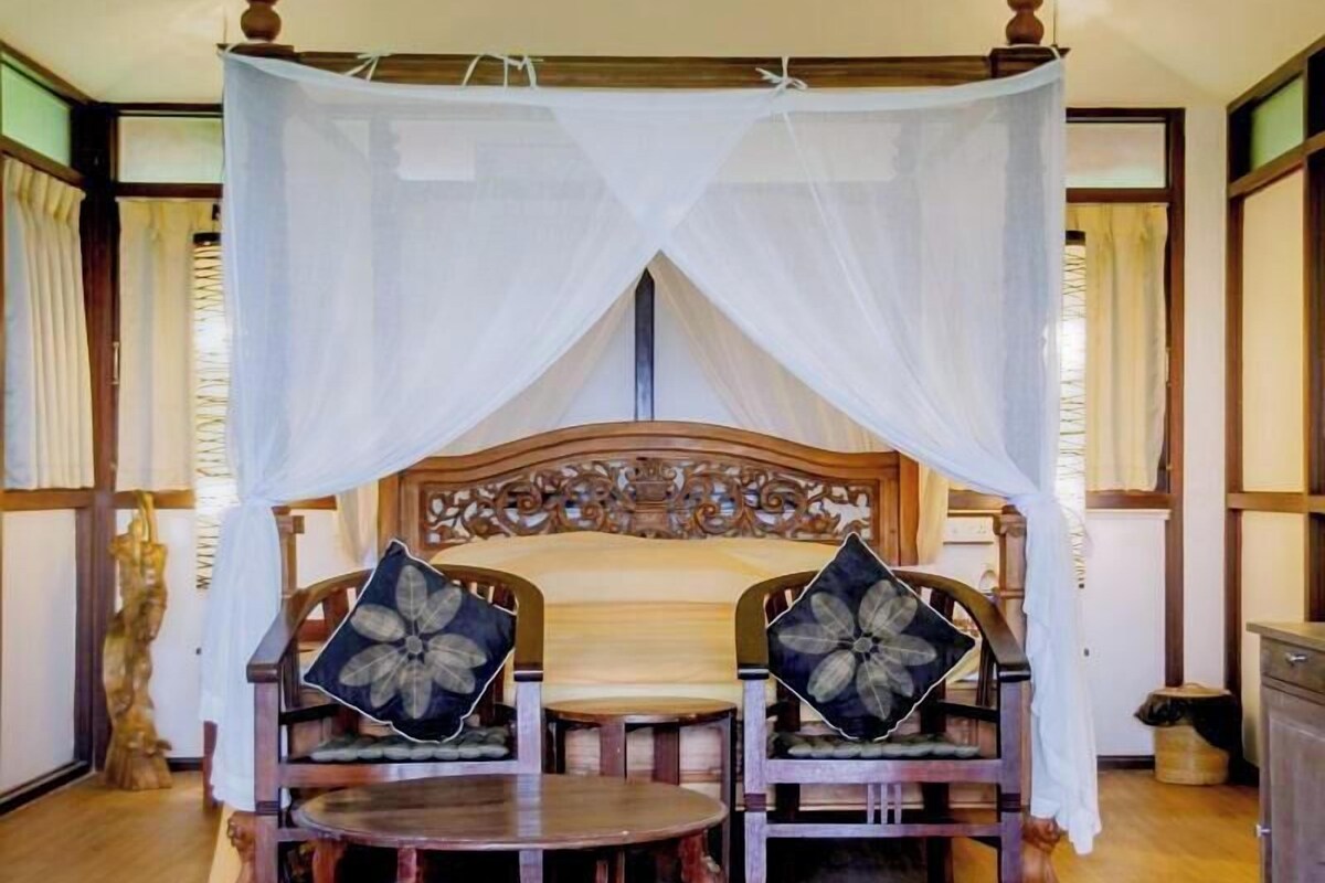 Quarto com cama de dossel, duas cadeiras com almofadas florais, cabeceira de madeira e uma pequena mesa redonda.