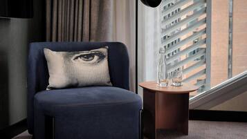 Quarto moderno com poltrona azul, uma pequena mesa de madeira com um copo e uma garrafa, e vista de um edifício por uma janela com persianas.