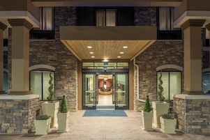 Entrada de hotel com fachada de pedra, teto de madeira e porta de vidro que dá acesso ao interior.