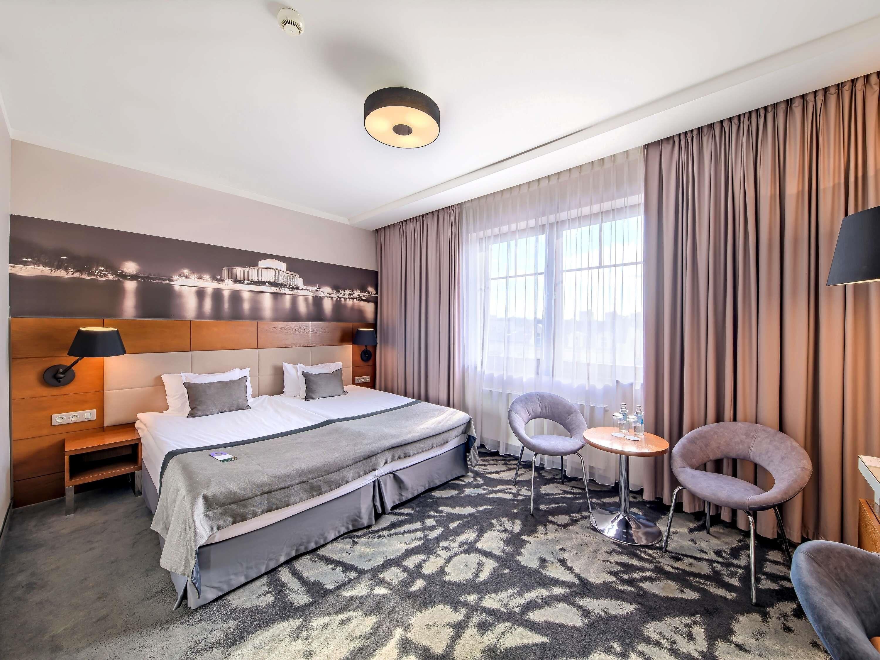 Quarto de hotel com cama, mesas de cabeceira, cabeceira com mural da paisagem urbana, pequena mesa redonda com duas cadeiras e tapete estampado.