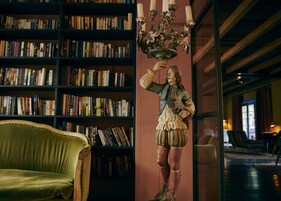 Uma biblioteca com estantes, uma poltrona verde e uma estátua de uma figura histórica.