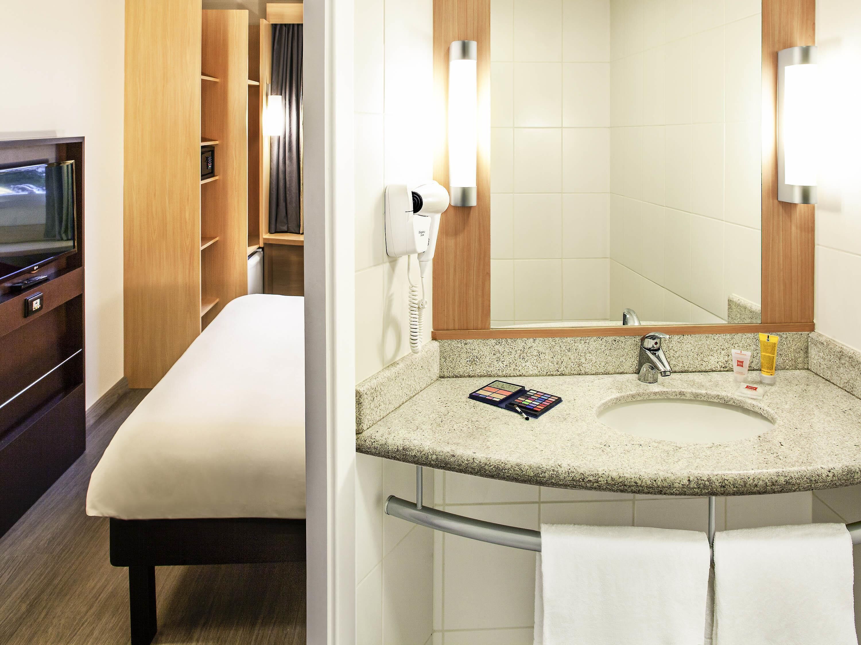 Quarto de hotel com uma cama, televisão, banheiro com pia, espelho e secador de cabelo.