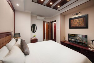 Quarto de hotel com uma cama grande, uma televisão exibindo 'Girlboss', uma obra de arte emoldurada na parede e uma cabeceira de madeira.