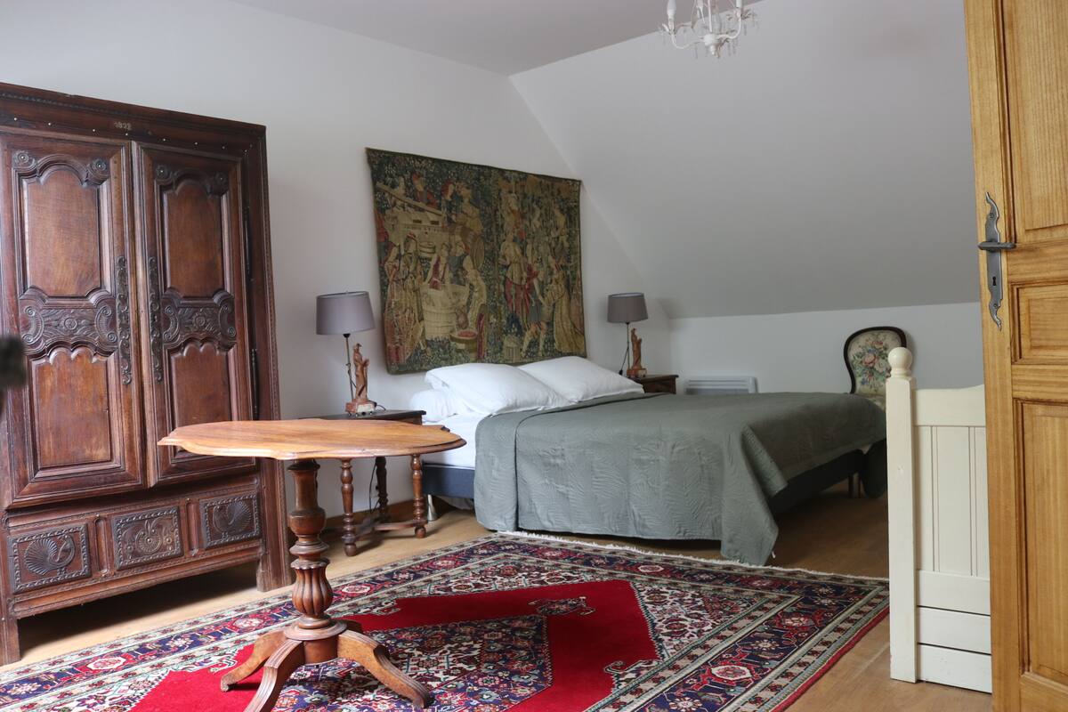 Quarto com um grande guarda-roupa de madeira, uma mesa redonda de madeira, uma cama com colcha cinza, tapeçaria na parede e um tapete estampado vermelho.