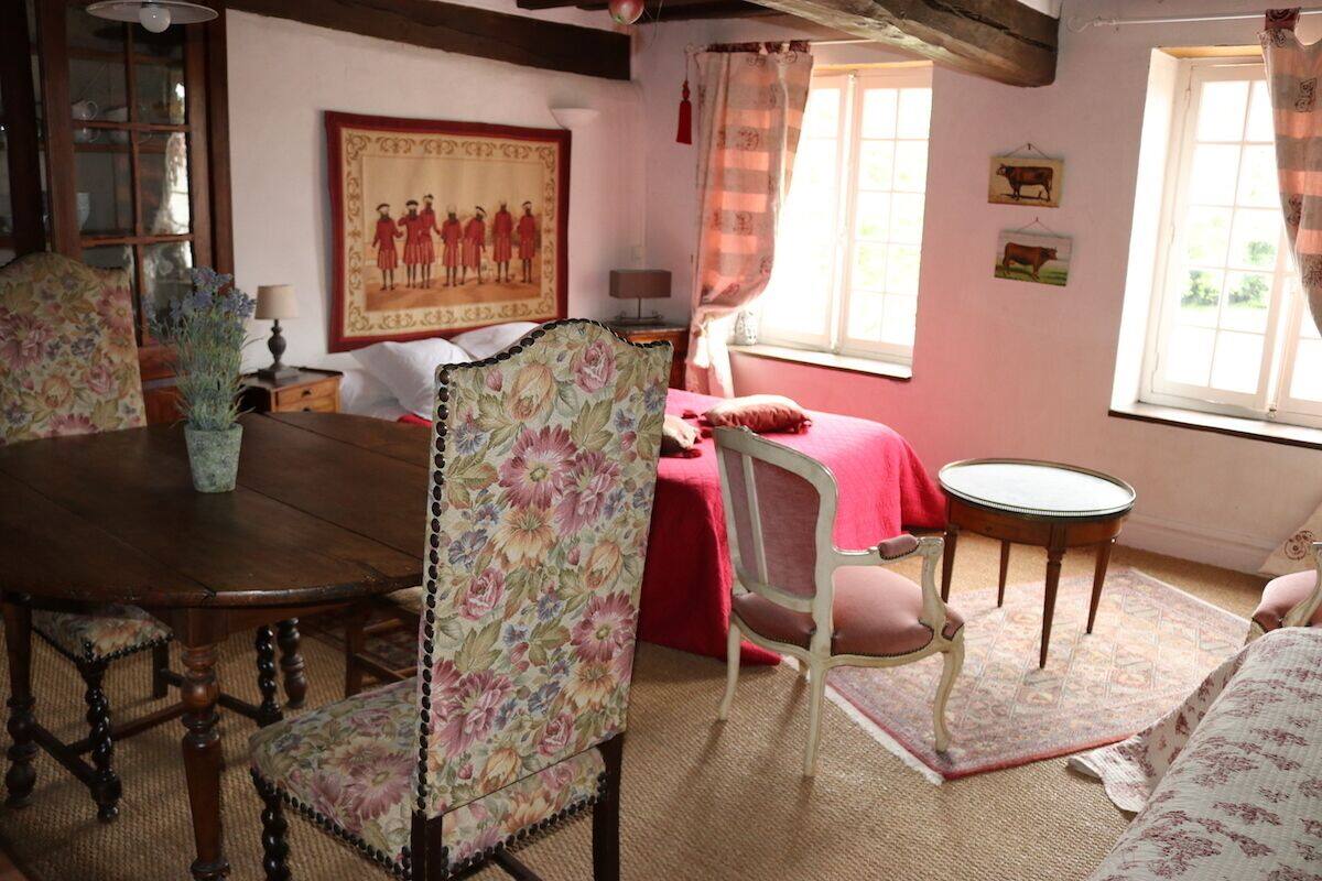 Quarto tradicional com mesa de jantar de madeira, cadeiras estofadas com motivos florais, cama vermelha e janela com cortinas.