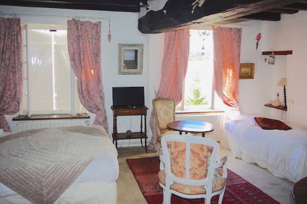 Quarto com cama, cadeira, mesinha, televisão e janela com cortinas.