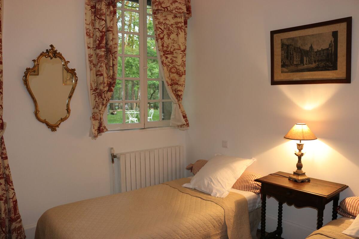 Um quarto com cama, mesa de cabeceira com abajur, janela com cortinas, espelho e um quadro na parede.