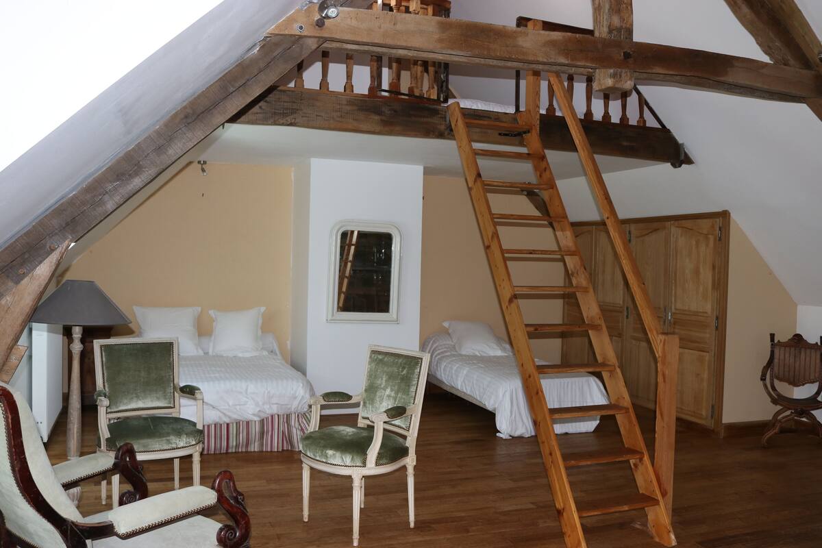 Um quarto em estilo loft com escada de madeira, duas poltronas estofadas verdes, uma cama de madeira e uma pequena mesa.