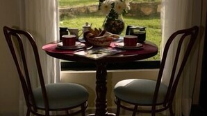 Uma sala de jantar com uma mesa e duas cadeiras, um vaso com flores e vista para um jardim pela janela.