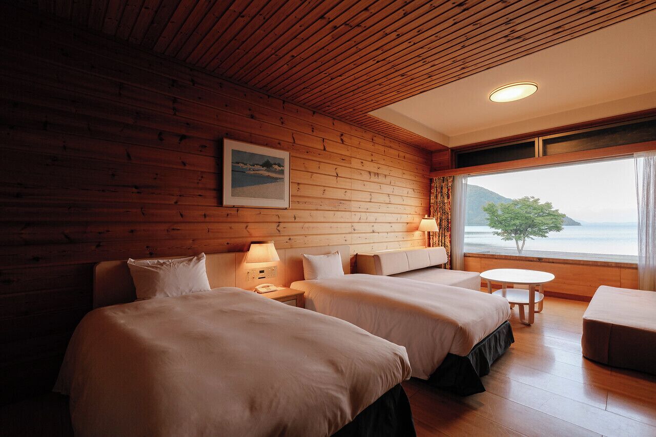 Quarto de hotel com duas camas, uma mesinha e janela com vista para o mar.
