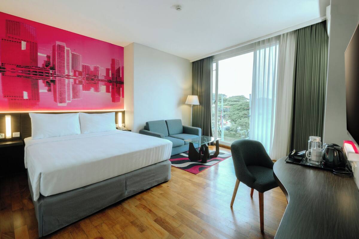 Quarto de hotel com cama grande, sofá, escrivaninha e um mural com vista para a cidade.