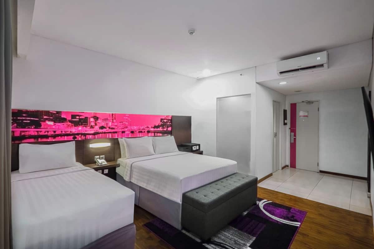 Quarto de hotel com duas camas, uma televisão grande fixada na parede, um aparelho de ar condicionado e uma porta com detalhe vermelho.