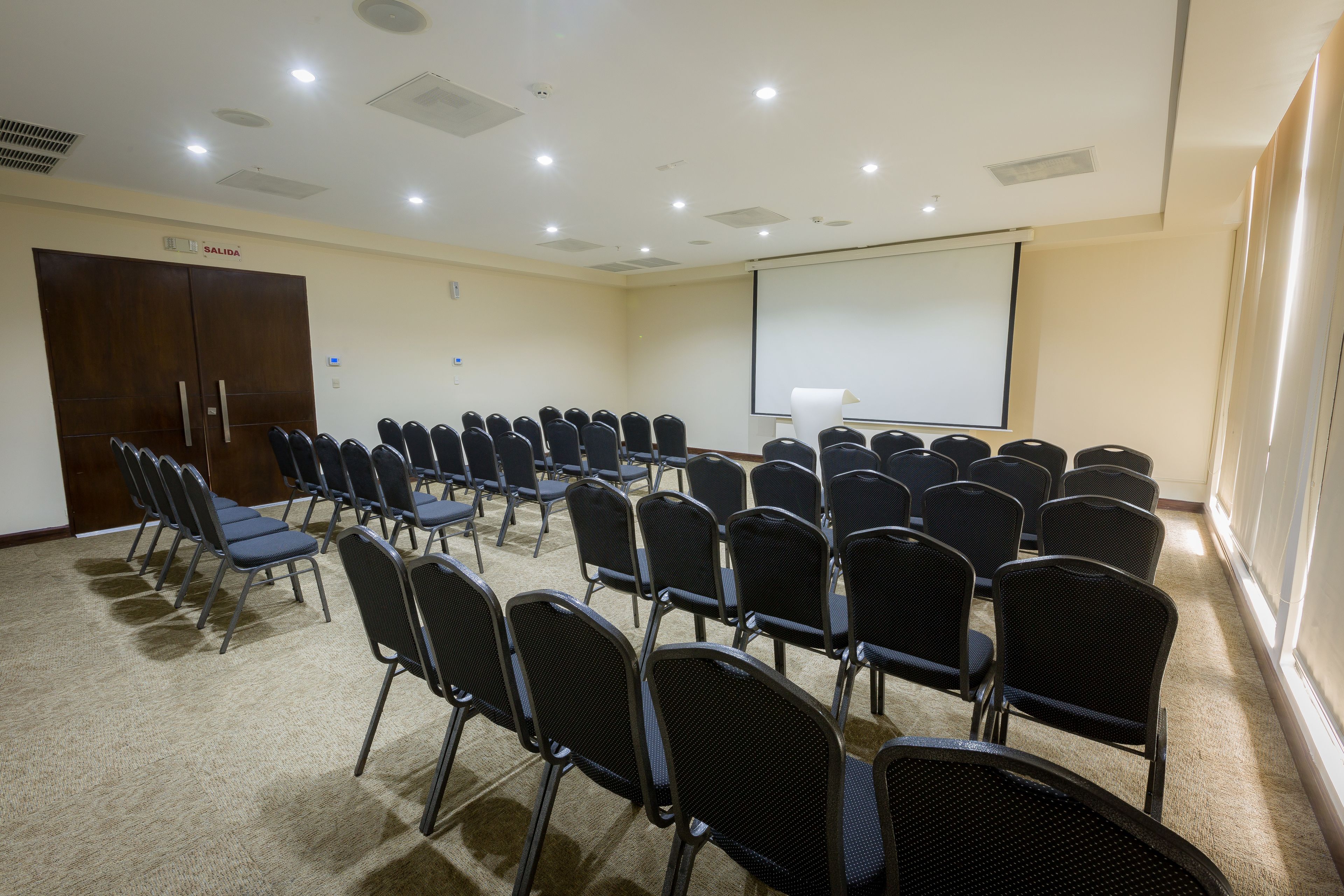 Uma sala de conferências com fileiras de cadeiras, uma tela de projetor e uma porta identificada com a palavra 'SALIDA'.
