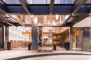 Um lobby moderno de hotel com fachada de vidro, escadaria central e balcão de recepção.