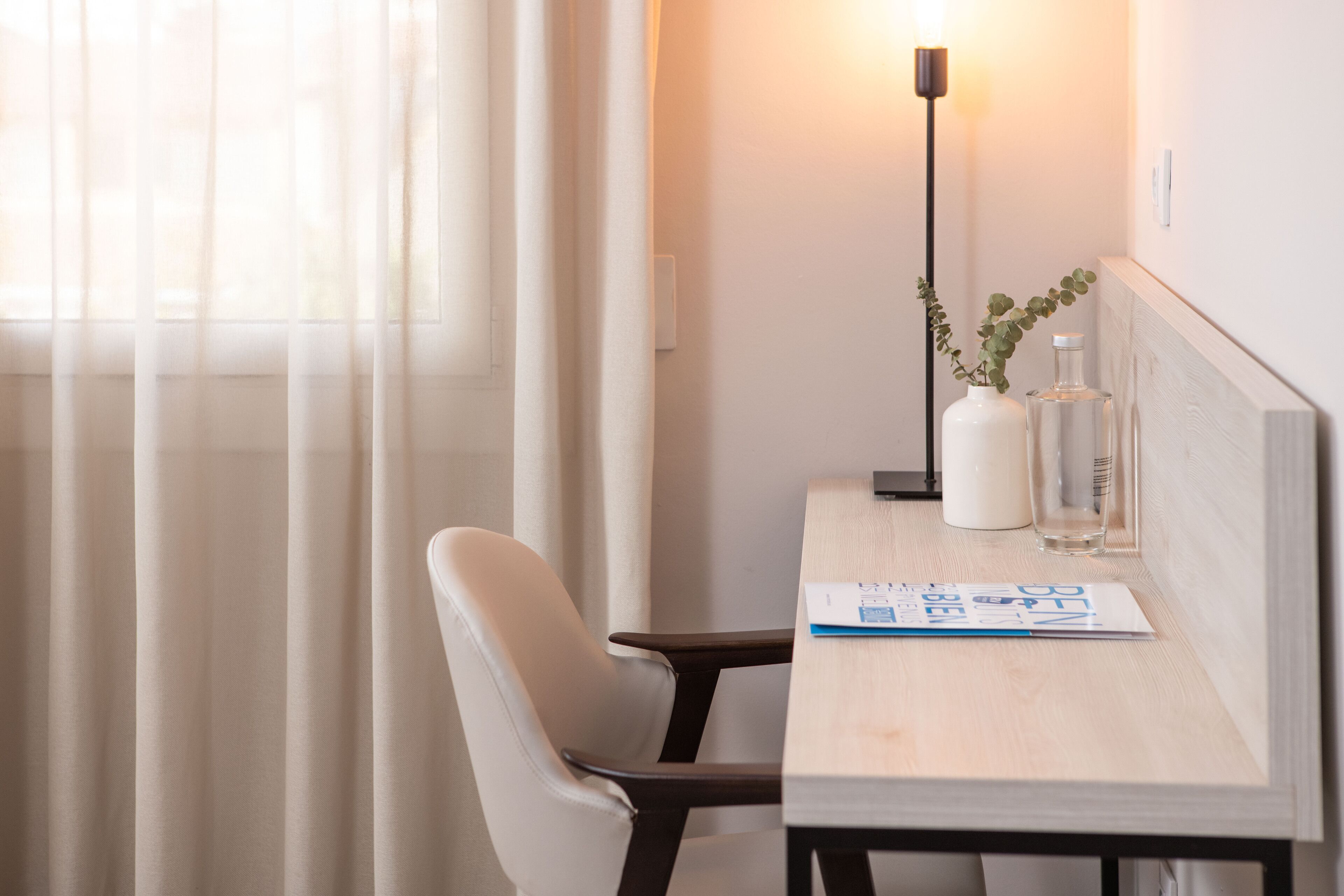 Quarto de hotel com uma escrivaninha de madeira, uma cadeira, uma luminária, um vaso com uma planta e uma garrafa.