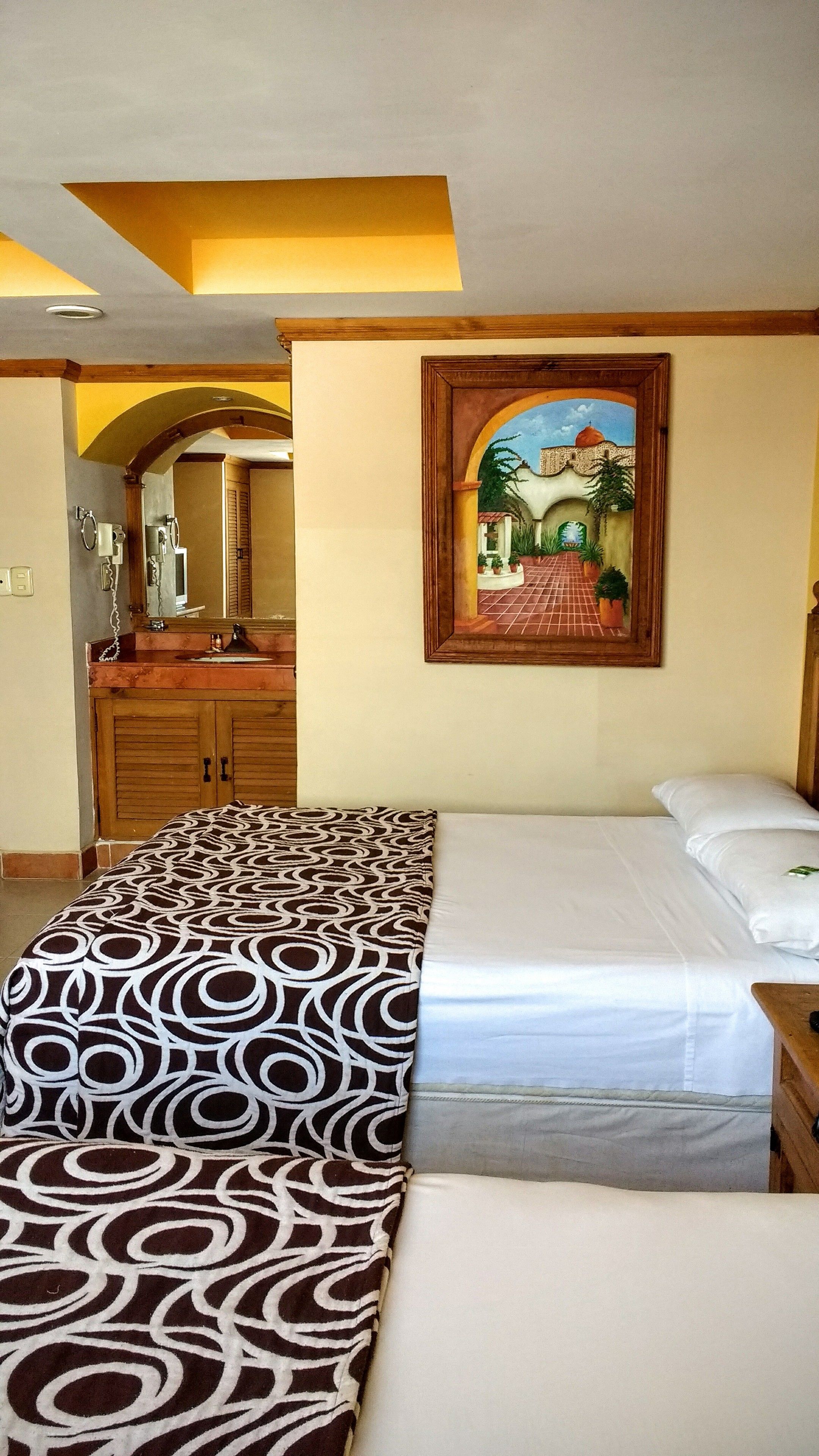 Quarto de hotel com cama, espelho grande, pintura de paisagem e toucador de madeira com pia.