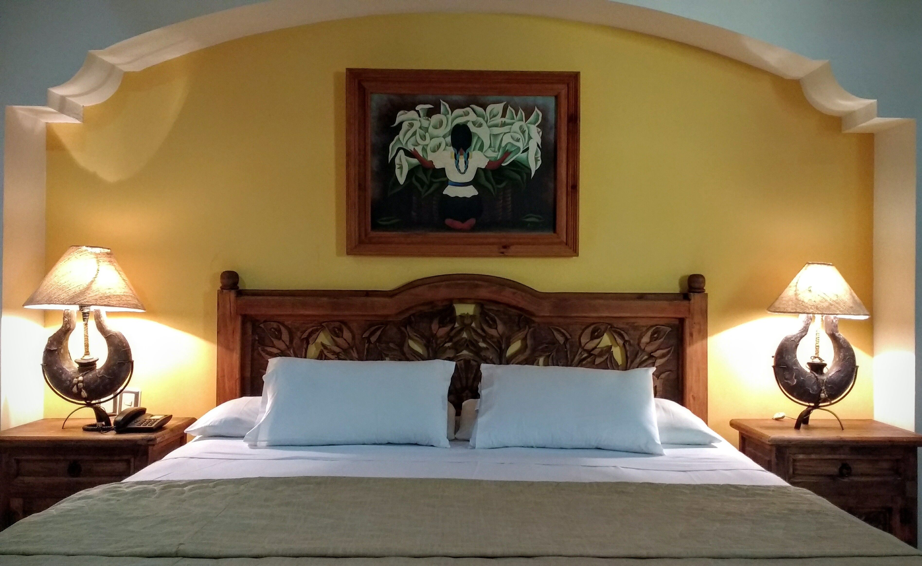 Quarto de hotel com cabeceira de madeira, duas luminárias de cabeceira, cama com roupa de cama branca e um quadro emoldurado acima da cabeceira.