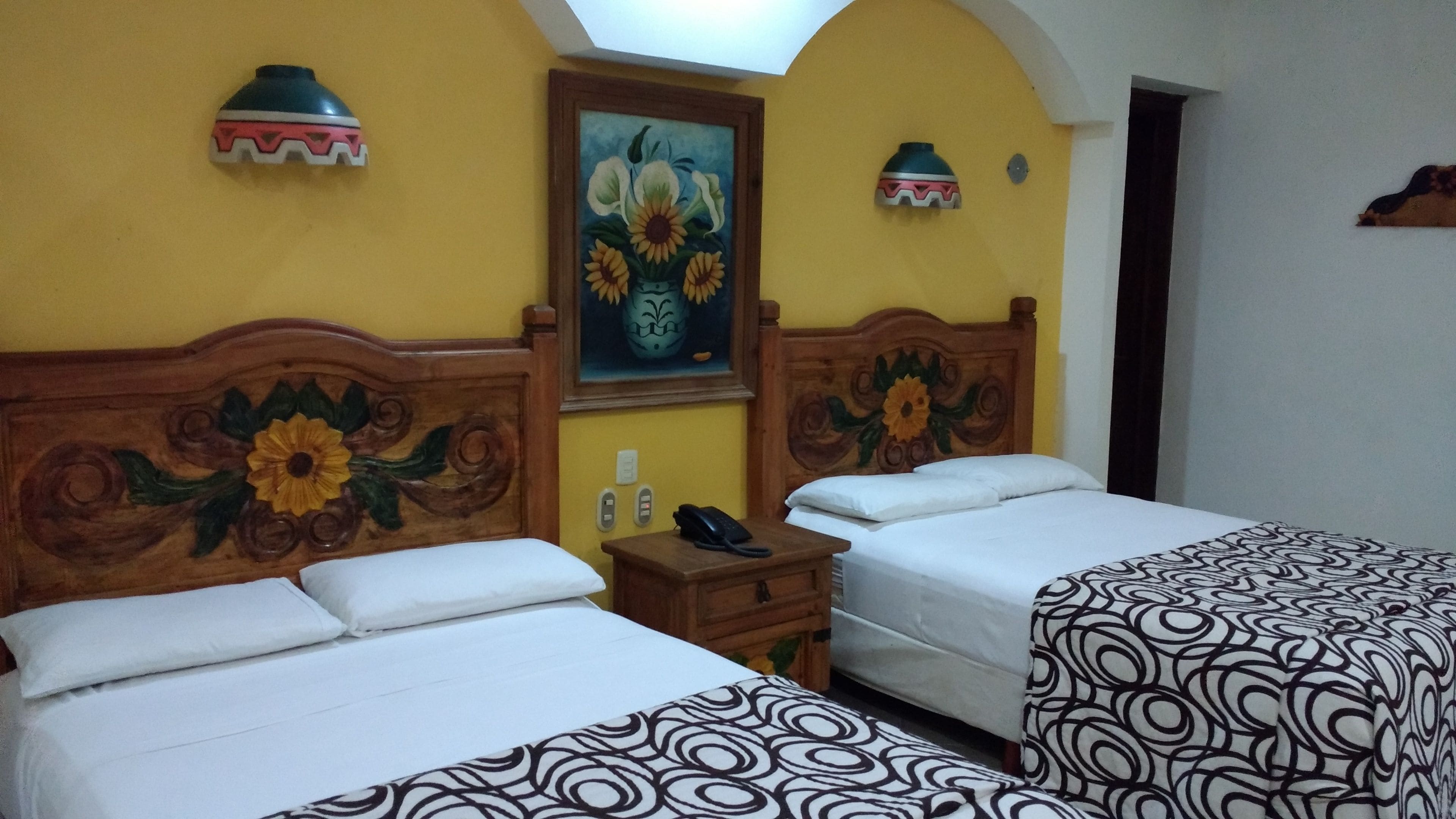 Quarto de hotel com duas camas, cabeceira de madeira com detalhes florais, pintura de girassóis, mesa de cabeceira de madeira e telefone fixado na parede.