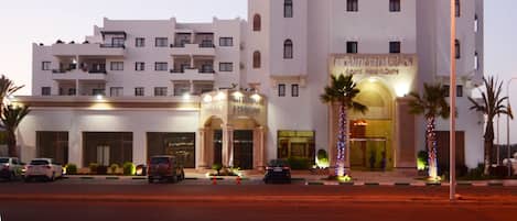 Um hotel com vários andares, uma entrada notável com arco e um estacionamento com diversos carros.