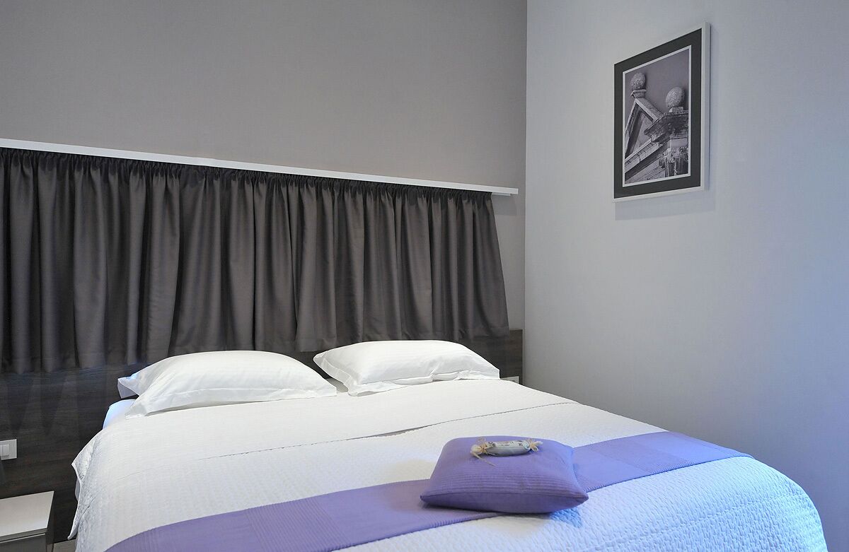 Uma cama bem arrumada com lençóis brancos e um travesseiro roxo, cortina cinza escura e um quadro na parede.