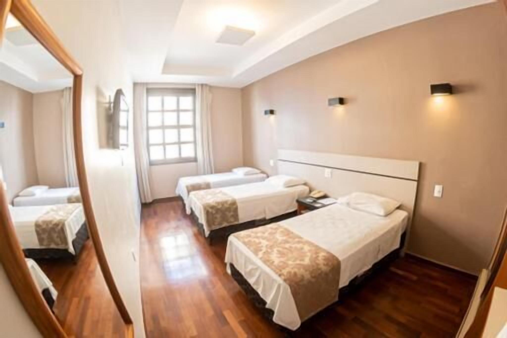 Quarto de hotel com três camas de solteiro, um espelho grande e uma janela com cortinas.
