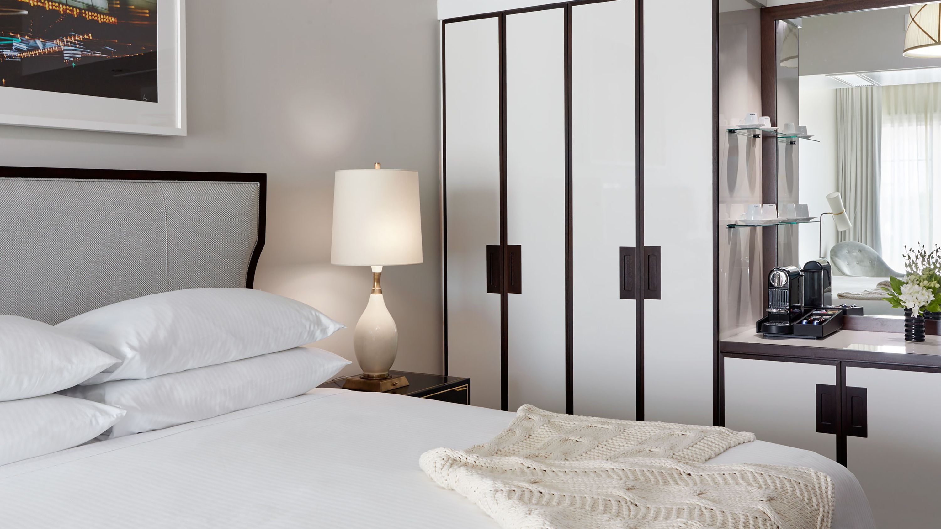 Uma cama bem arrumada com lençóis e almofadas brancos, uma luminária de cabeceira, um espelho e uma cafeteira em uma mesinha de cabeceira.