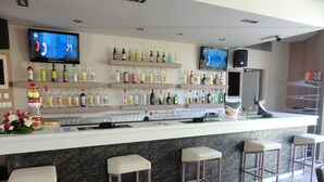 Uma área de bar com piso de mármore, diversas garrafas de bebidas nas prateleiras, banquetas e duas televisões fixadas na parede.