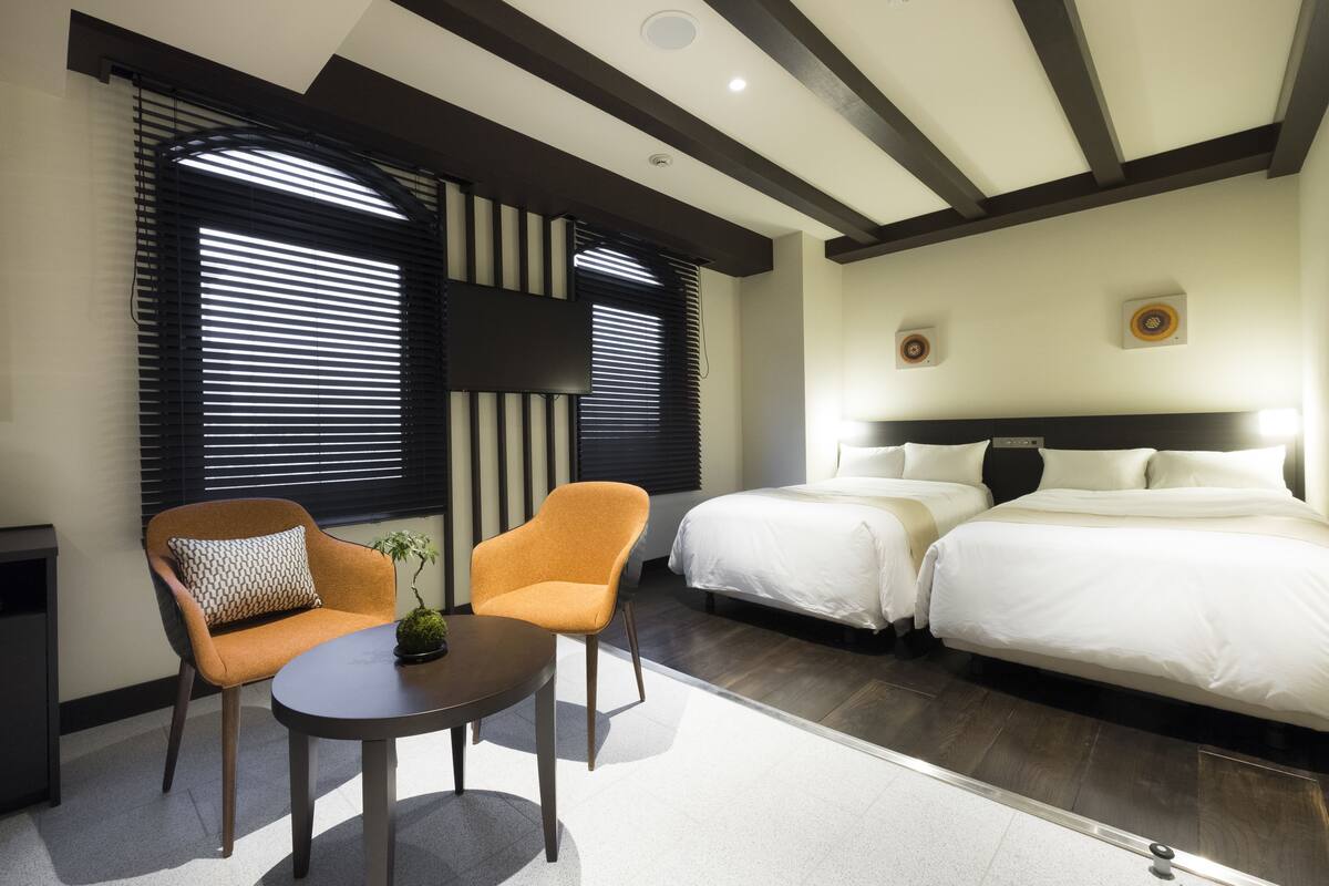 Quarto de hotel com uma cama, duas cadeiras, uma mesinha e um vaso de planta.