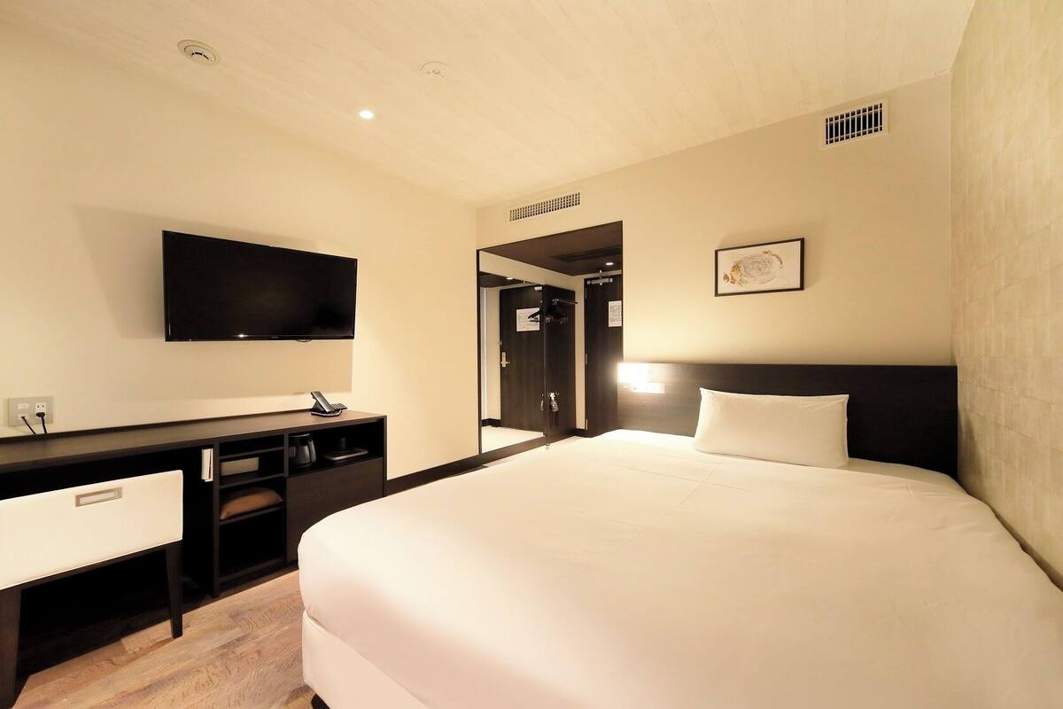 Quarto de hotel com cama, escrivaninha, cadeira, televisão e espelho.
