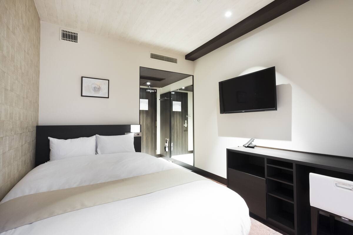 Quarto de hotel moderno com uma cama grande, televisão de tela plana na parede e uma escrivaninha embutida com espaço para armazenamento.