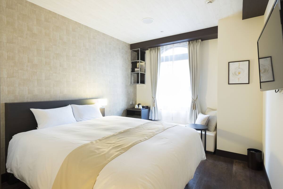 Quarto de hotel com uma cama grande, uma mesa de cabeceira, uma cadeira, uma pequena mesa, uma estante e uma janela com cortinas.
