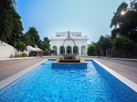 Uma piscina com uma fonte no centro, rodeada por um edifício branco com arcos verdes e uma cerca branca.
