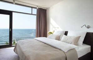 Quarto com uma janela ampla com vista para o mar, cama com roupa de cama branca e mesa de cabeceira com abajur e flores.