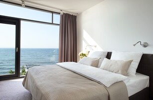Quarto com uma janela ampla com vista para o mar, cama com roupa de cama branca e mesa de cabeceira com abajur e flores.