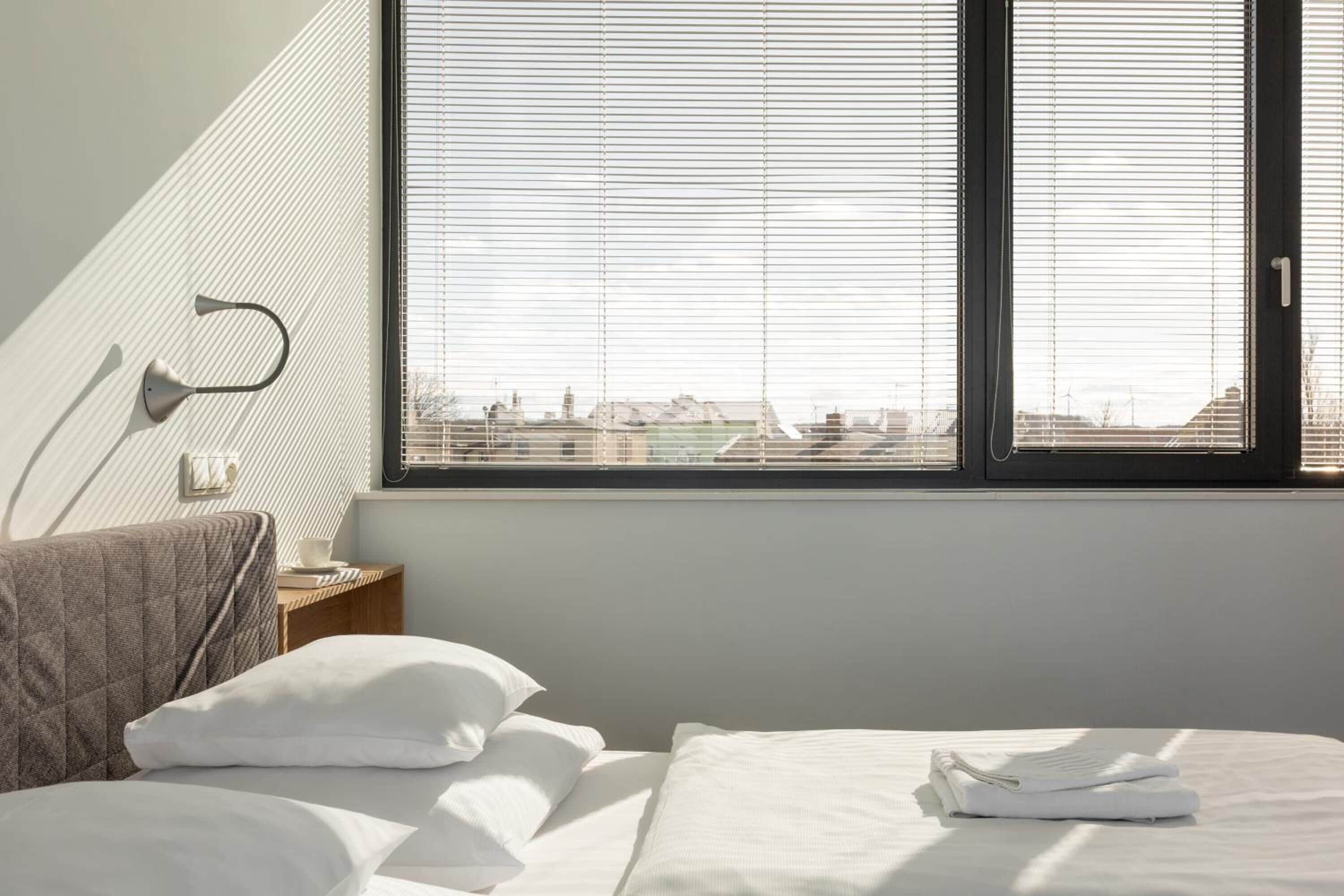Quarto de hotel com uma cama, roupa de cama branca, janela com persianas, abajur e vista para edifícios.