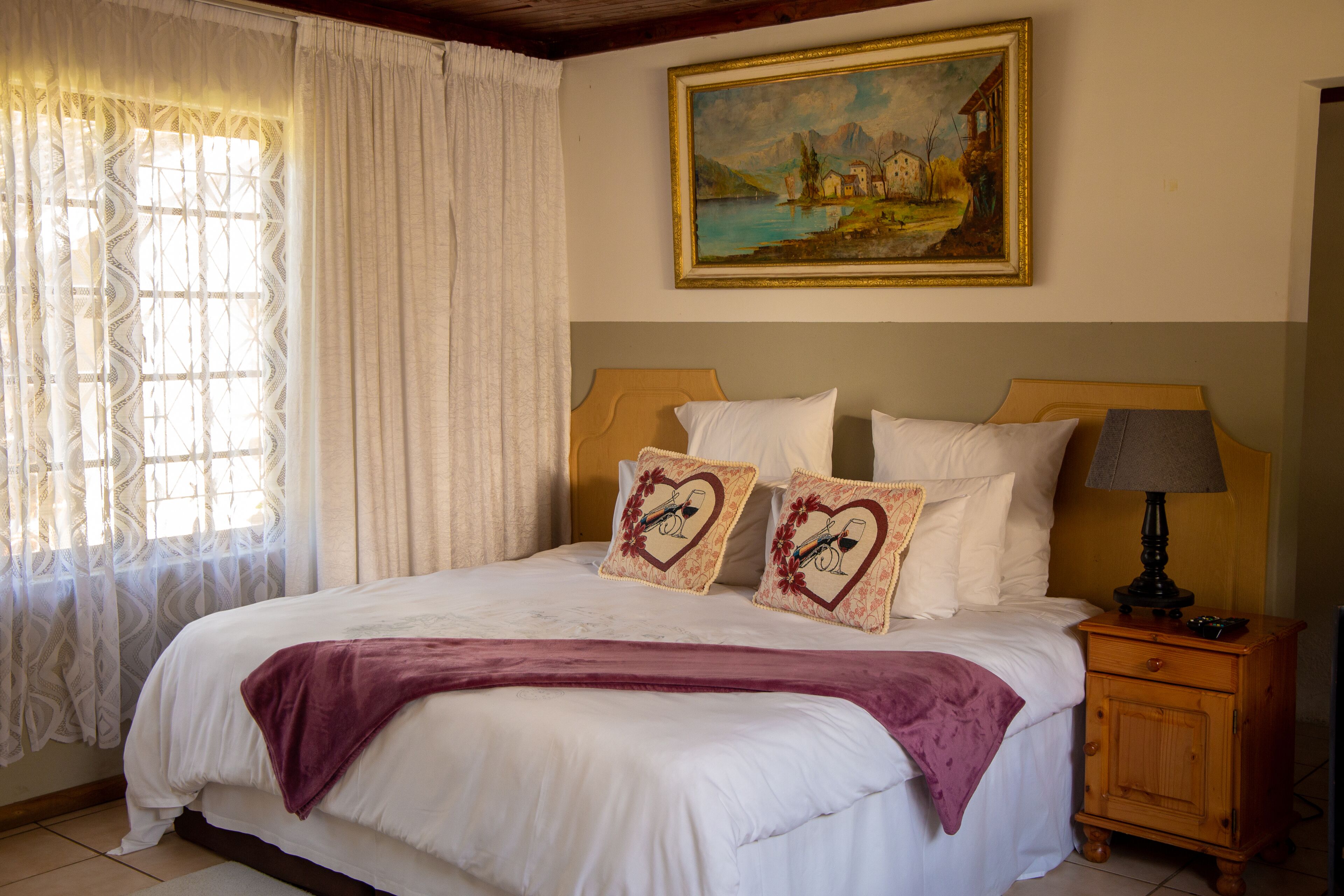 Uma cama bem arrumada com lençóis brancos e almofadas em formato de coração, uma mesa de cabeceira de madeira com uma luminária e um quadro de uma paisagem à beira de um lago na parede.