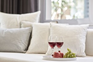 Uma cama com roupa de cama branca, dois copos de vinho tinto e um prato com uvas e uma fruta sobre um tabuleiro branco.