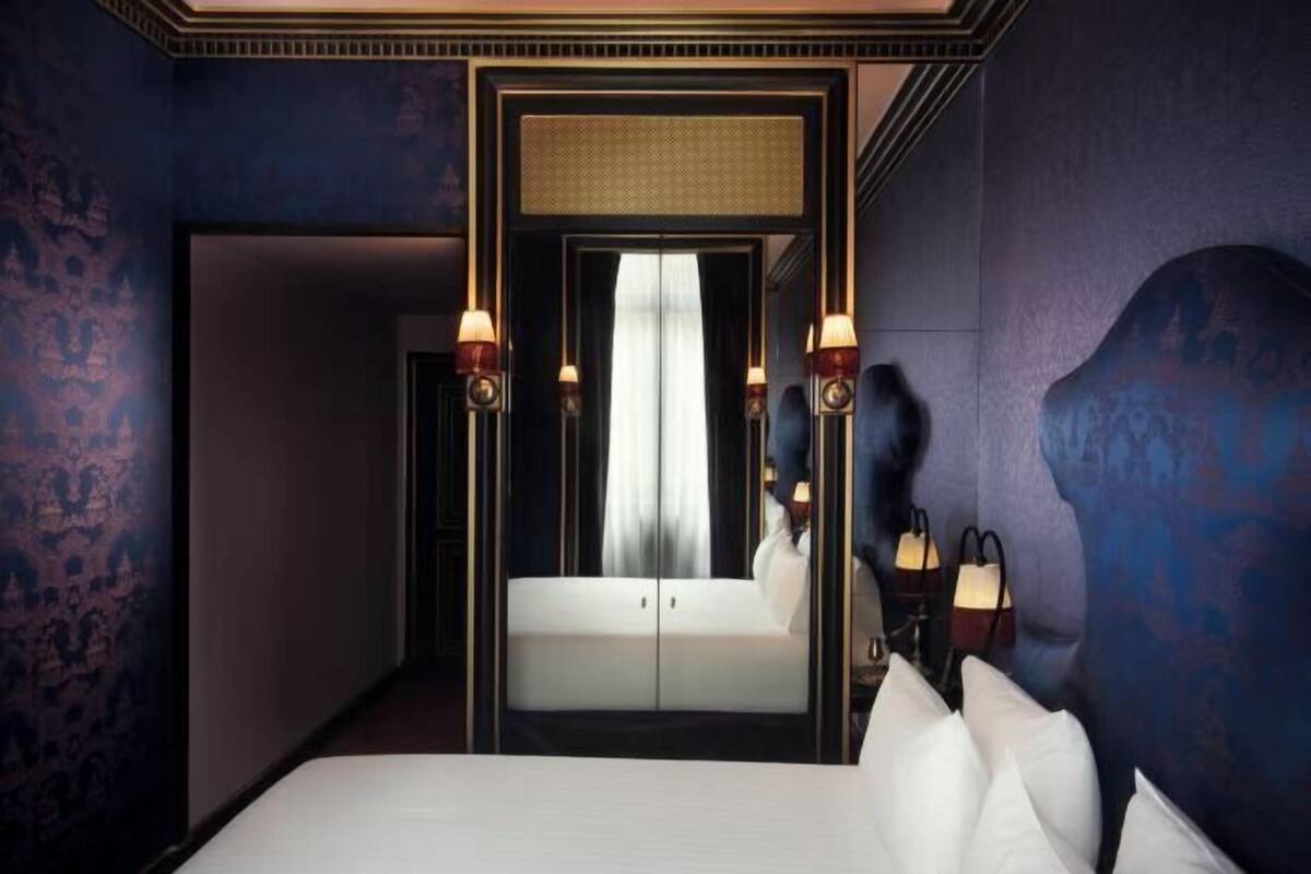 Quarto de hotel com uma cama grande, um espelho, duas luminárias fixadas na parede e vista para um sofá branco através de uma porta.