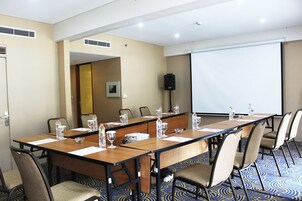 Uma sala de conferências com uma mesa comprida, cadeiras, uma tela de projetor e um palestrante.