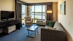 Quarto de hotel com televisão de tela plana, sofá, poltrona, mesa de centro e uma grande janela com vista para um corpo d'água.