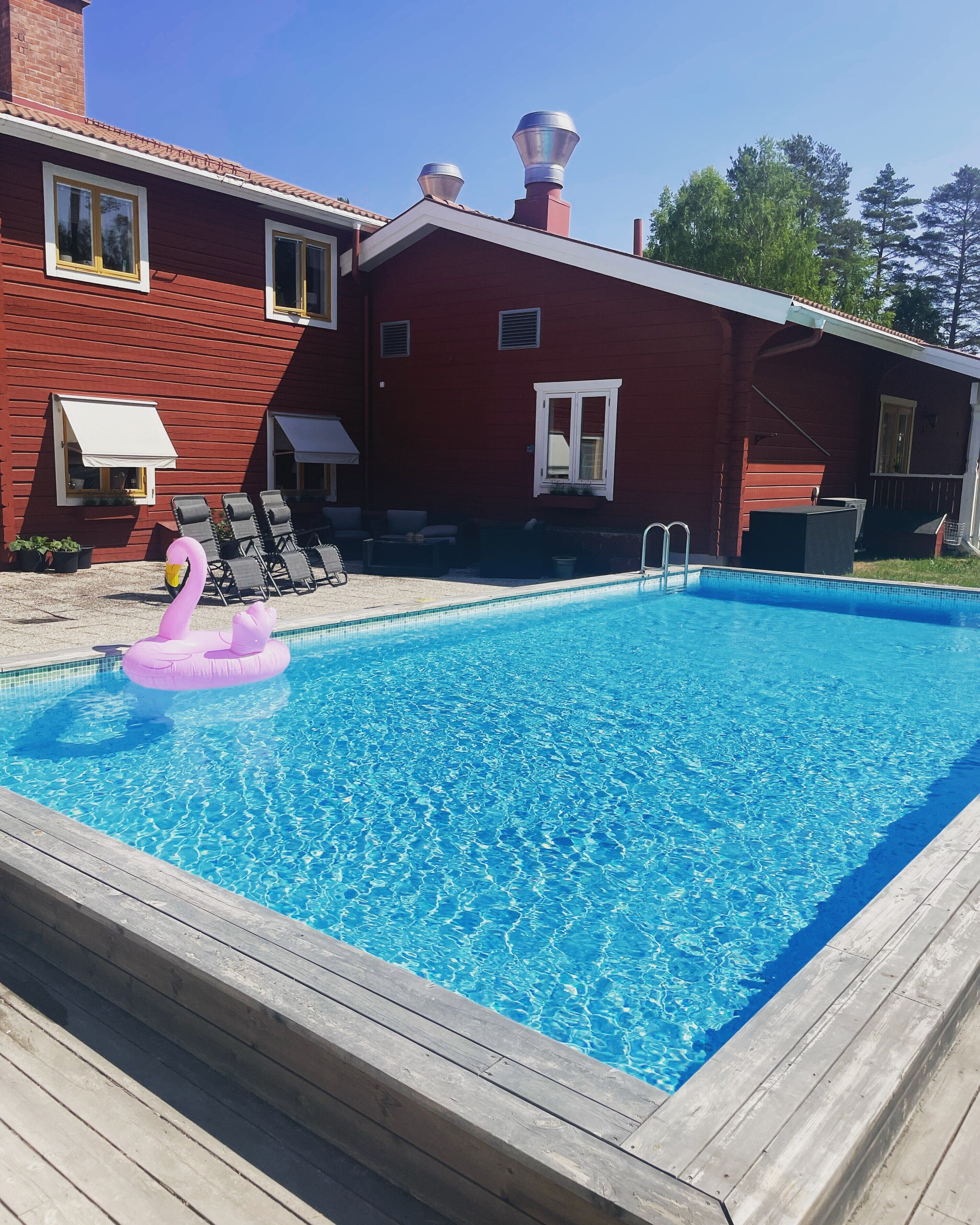 Uma piscina com um deck de madeira, um flutuador de flamingo rosa e um prédio vermelho com chaminé ao fundo.