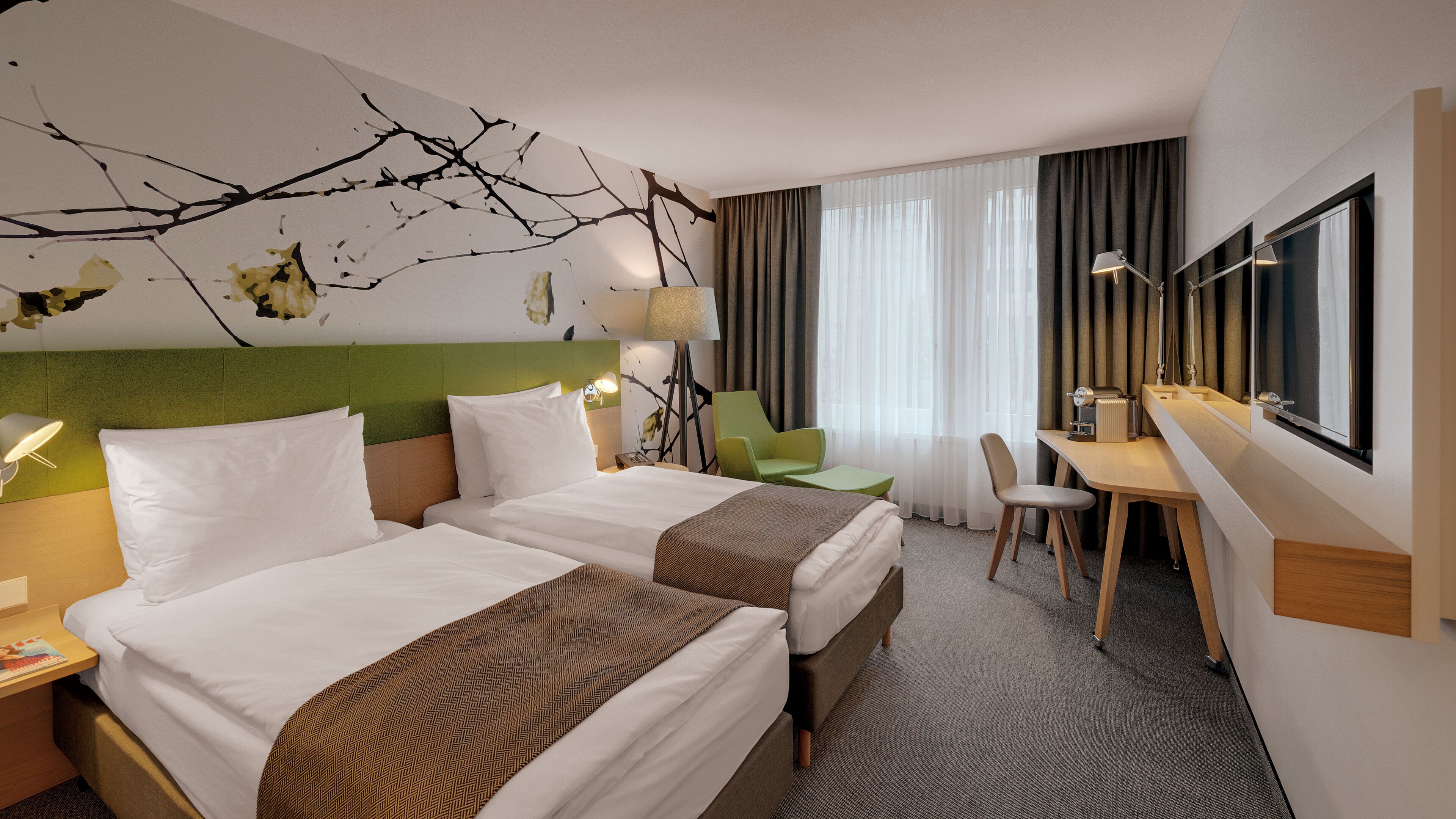 Quarto de hotel com duas camas, uma escrivaninha, uma cadeira, uma luminária e uma janela grande com cortinas.