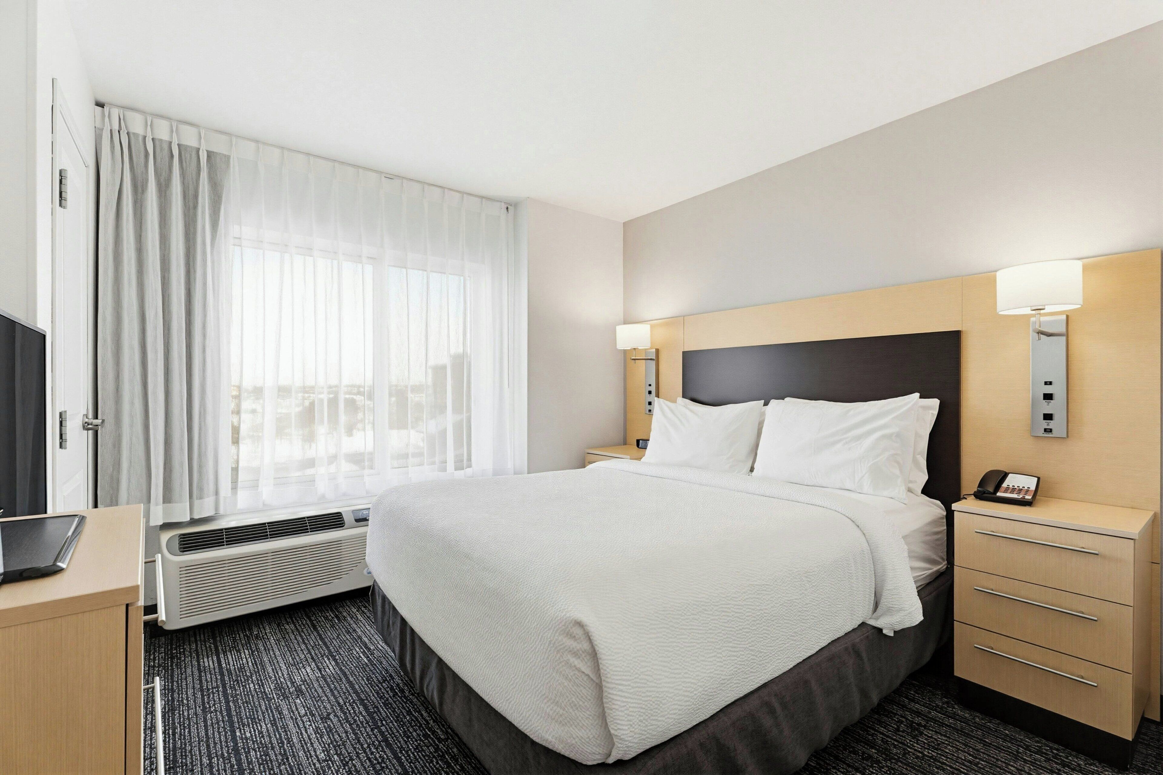 Quarto de hotel com cama, mesa de cabeceira, abajur e janela com cortinas.