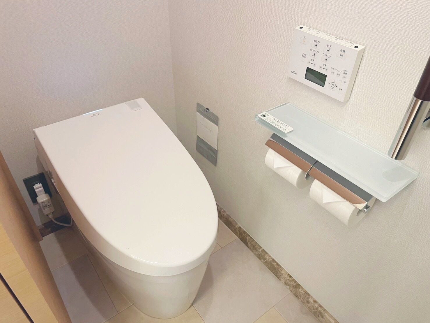Sanitário com botão de descarga, suporte para papel higiênico e painel de controle na parede.