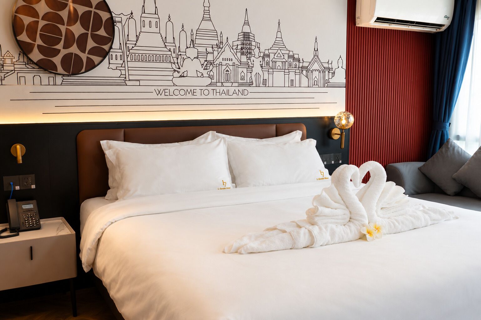 Quarto de hotel com cama, uma obra de arte na parede representando arquitetura tailandesa, arranjo de toalhas em forma de cisne e cortina vermelha.